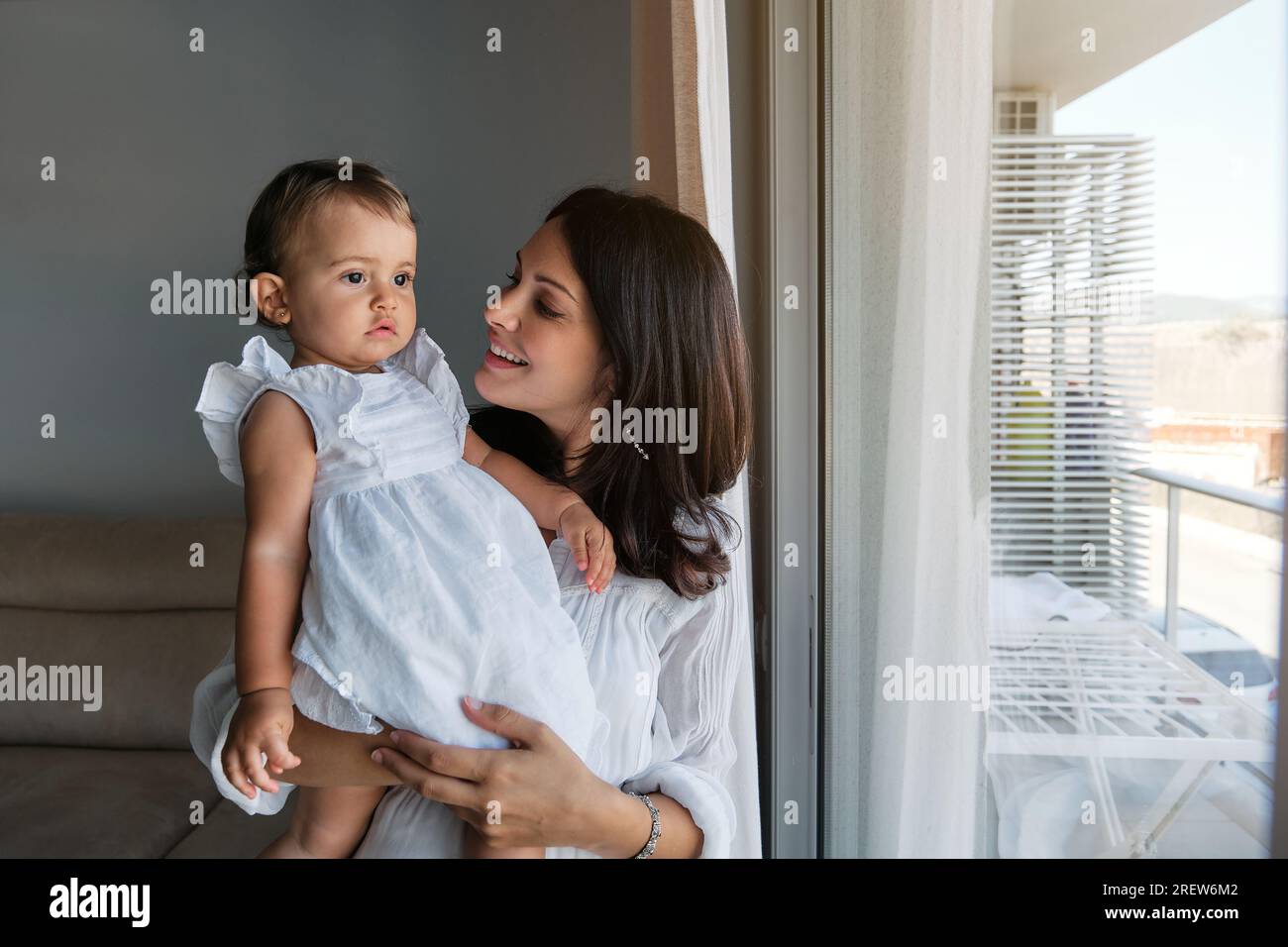 Curious baby in white dress looking out window with interest in arms of ...