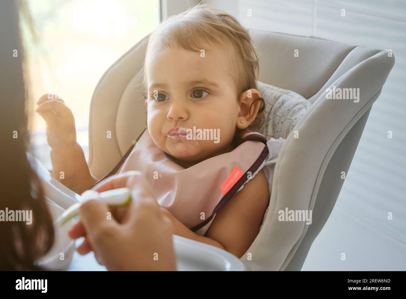 Adorable baby girl in bib sitting on high chair and looking at mother feeding her with spoon in ...