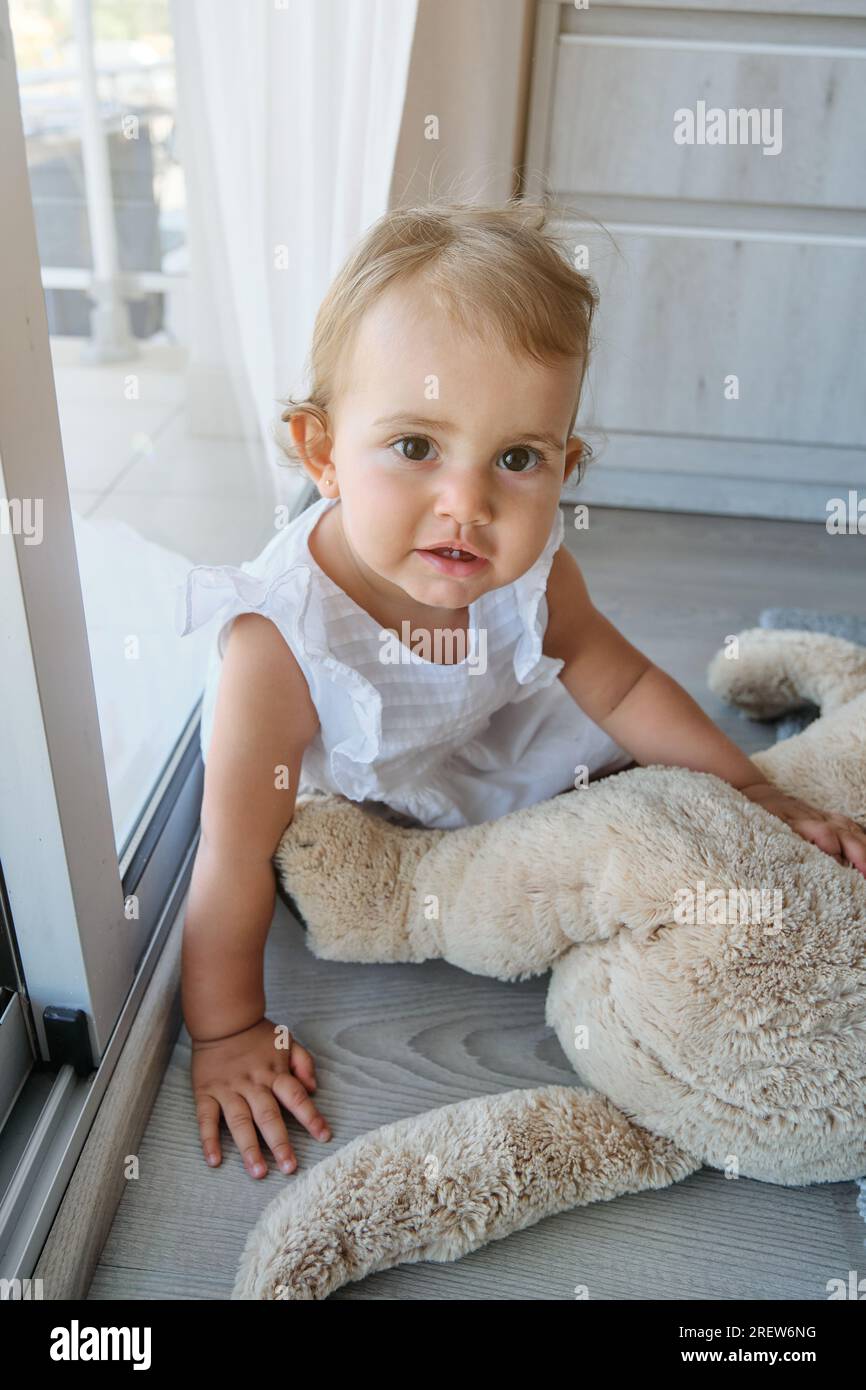 High angle of curious baby in white dress playing with plush toy and ...