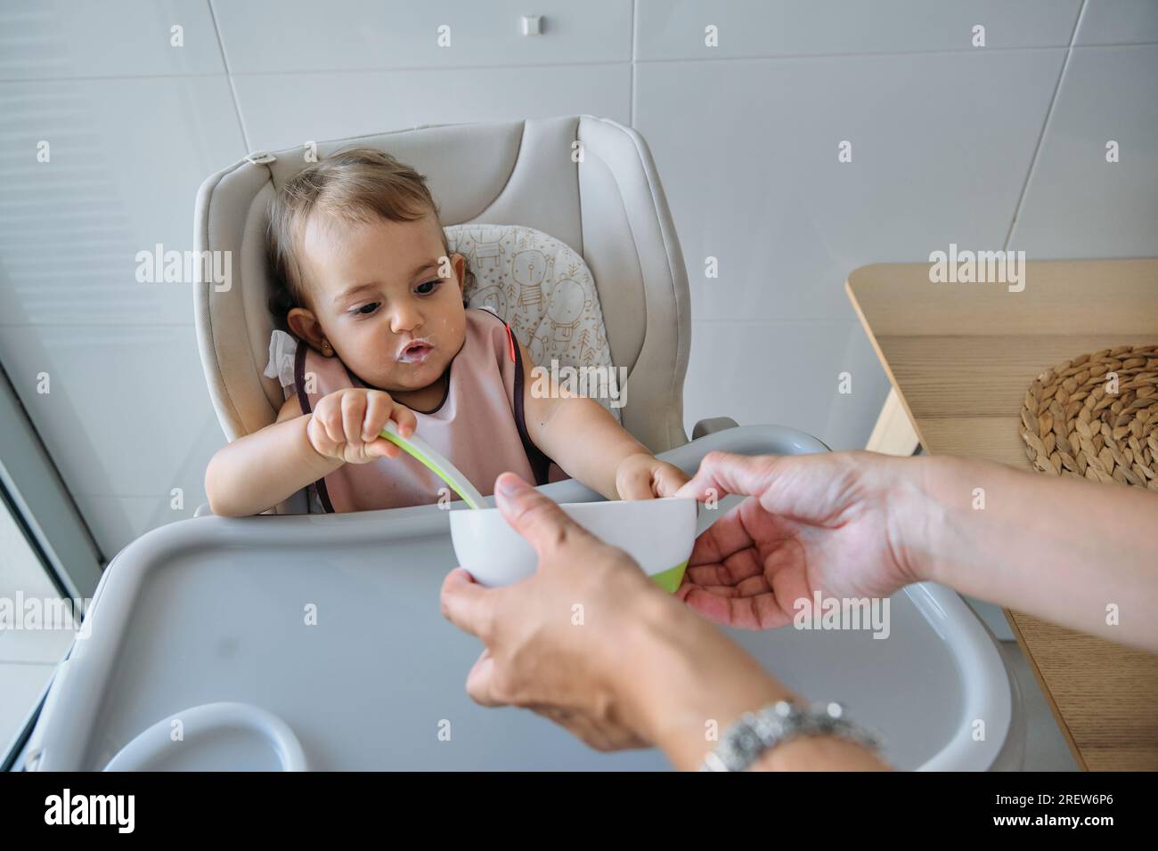 Adorable little girl in bib sitting on white high chair and eating with spoon while ...