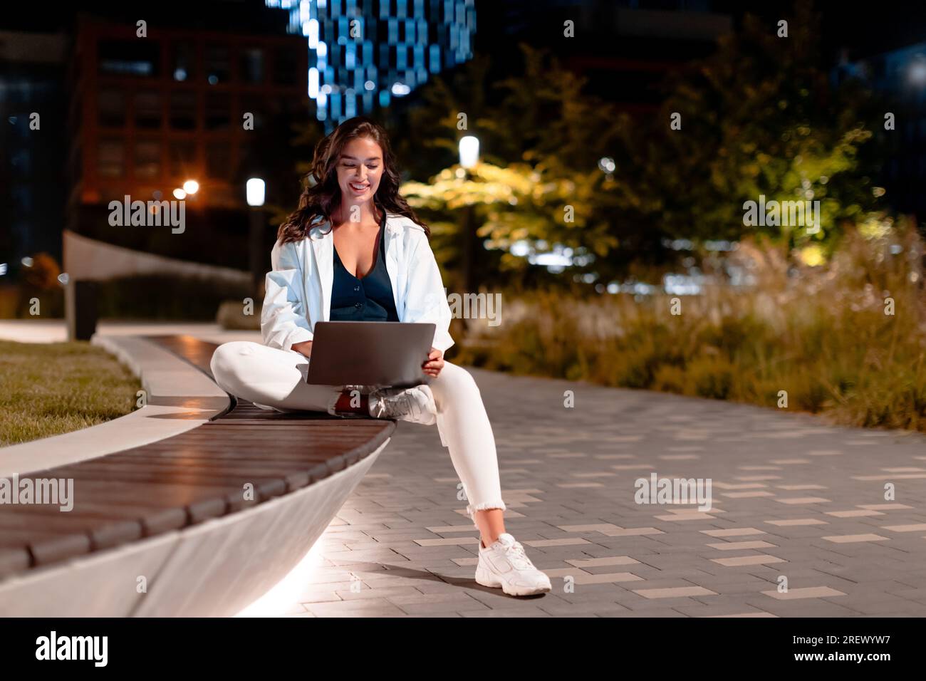 Female freelancer working on laptop, sitting on bench at night in city ...