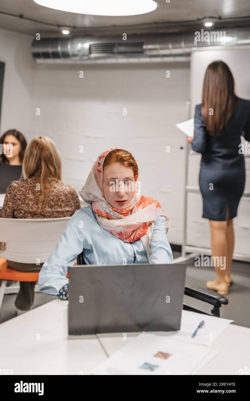 Disabled ginger female muslim employee in wheelchair working on lap top ...