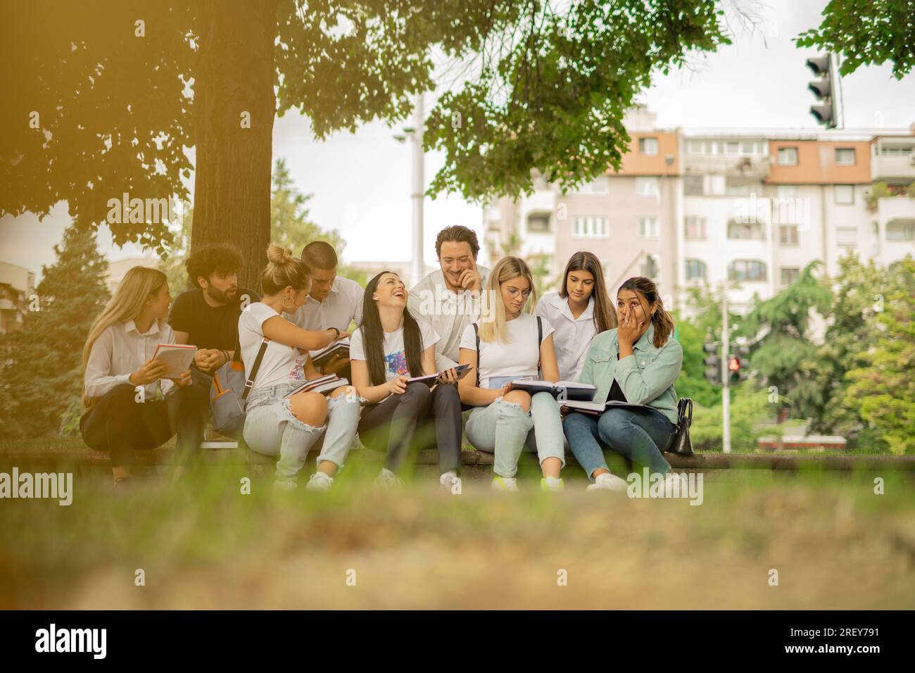 Young group of high school students sat together outside on the pathway with books in their ...