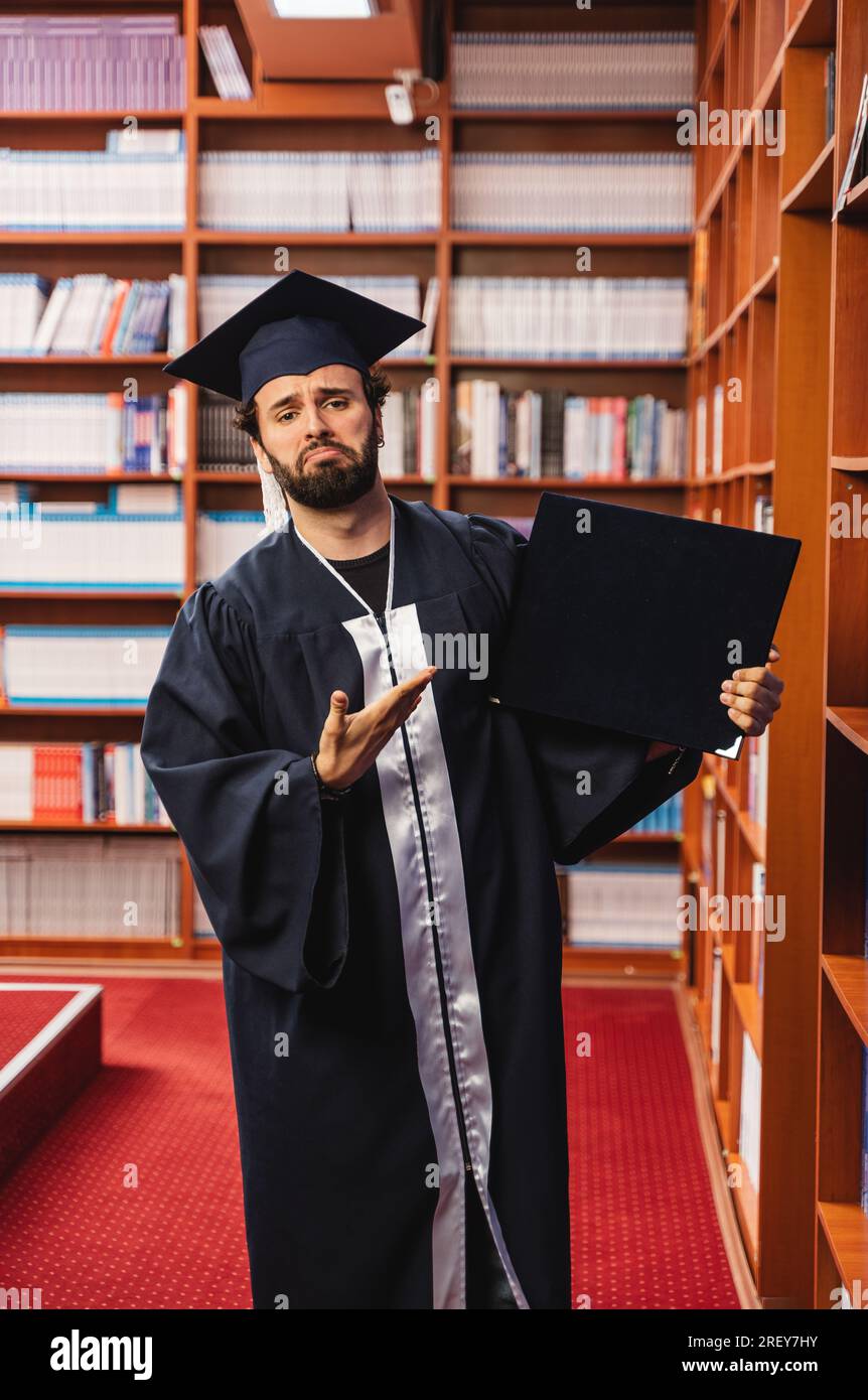 Young graduate wearing his cap and gown looking sad while holding his ...