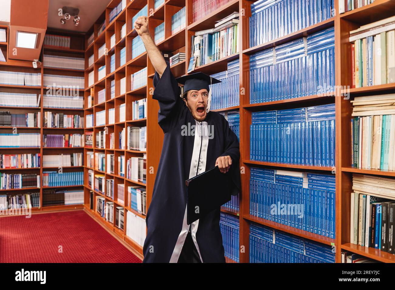 Shocked graduate wearing his cap and gown pumps his fist in the air and ...