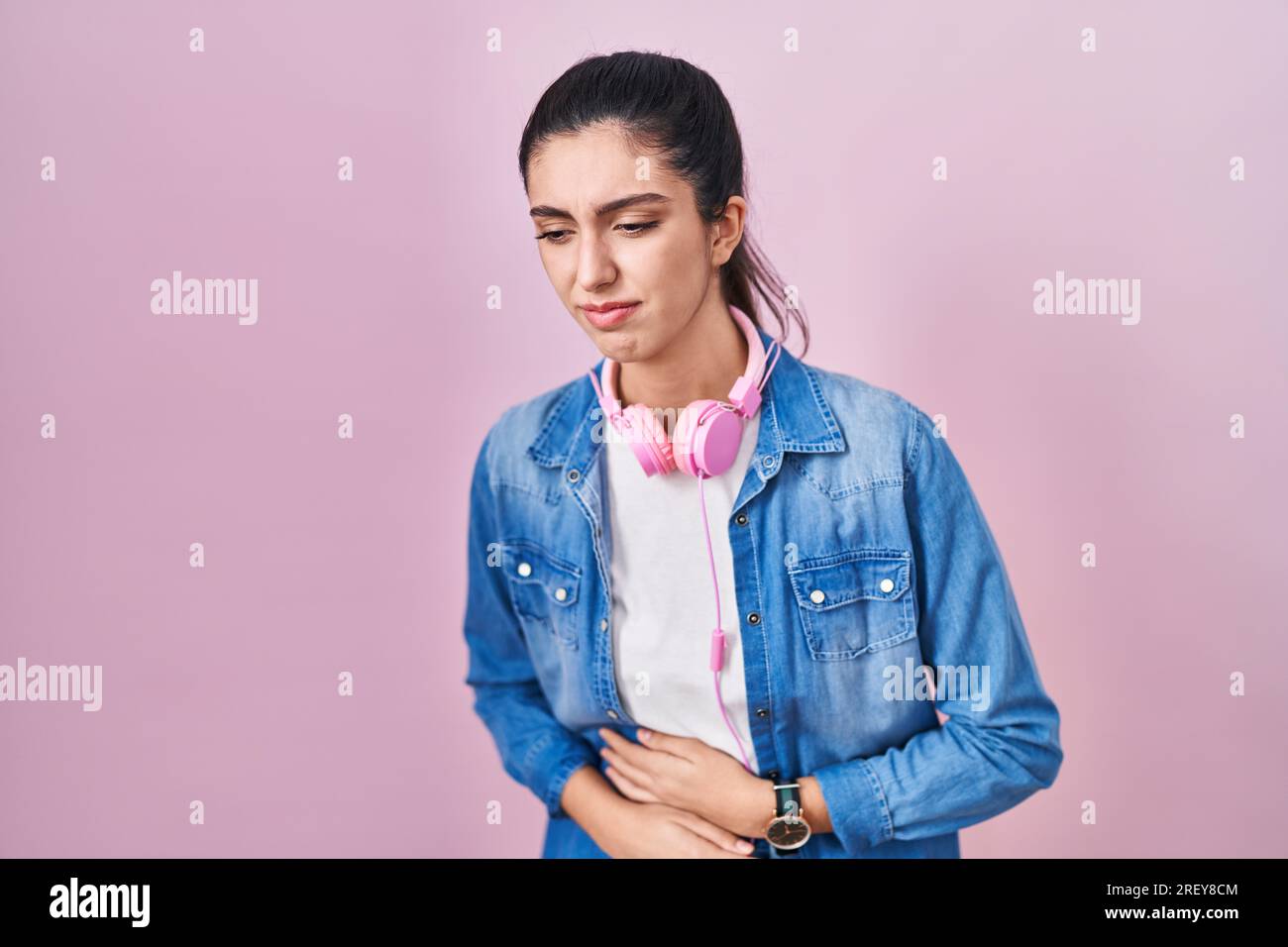Young beautiful woman standing over pink background with hand on stomach because nausea, painful ...
