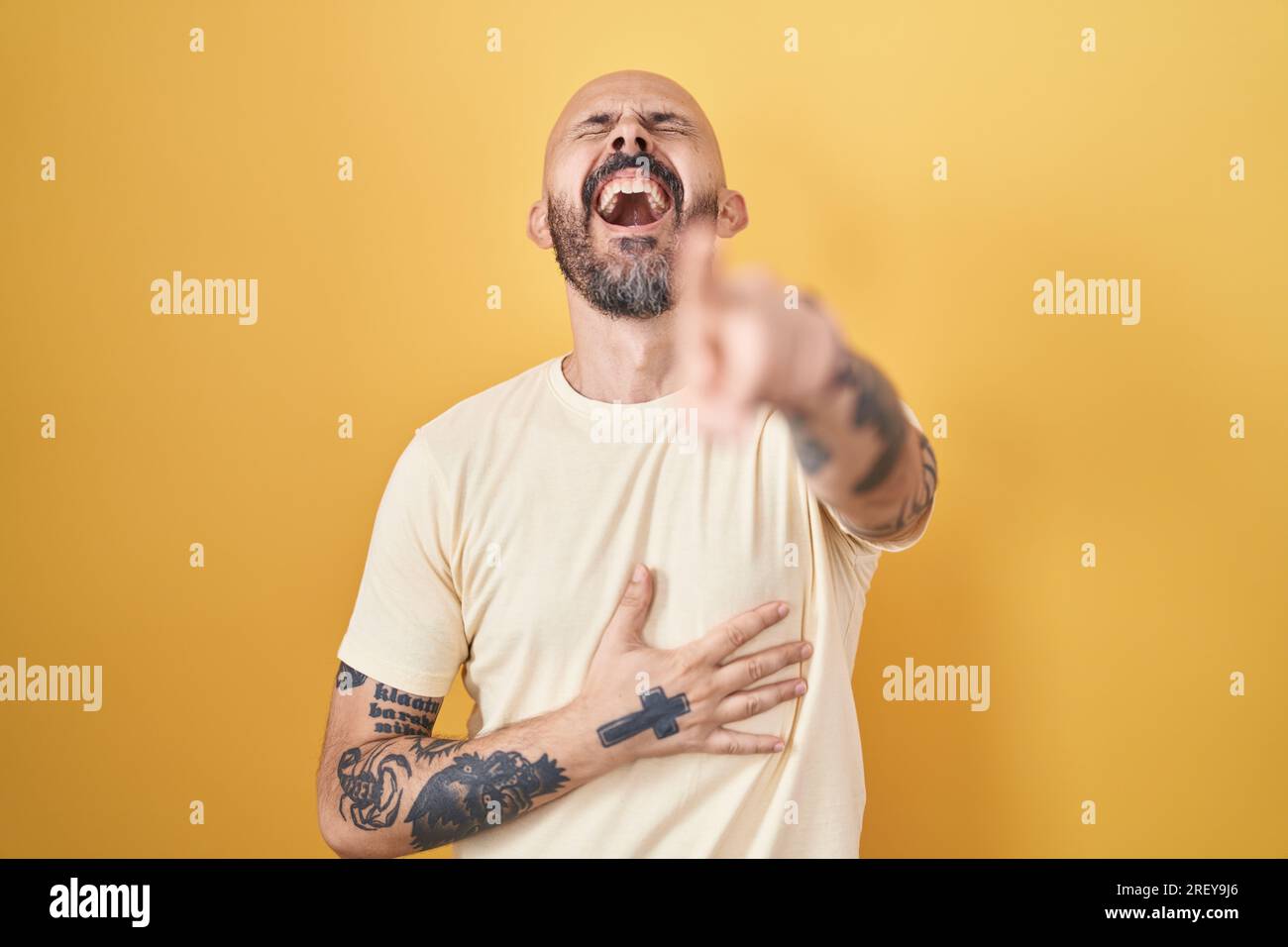 Hispanic man with tattoos standing over yellow background laughing at ...