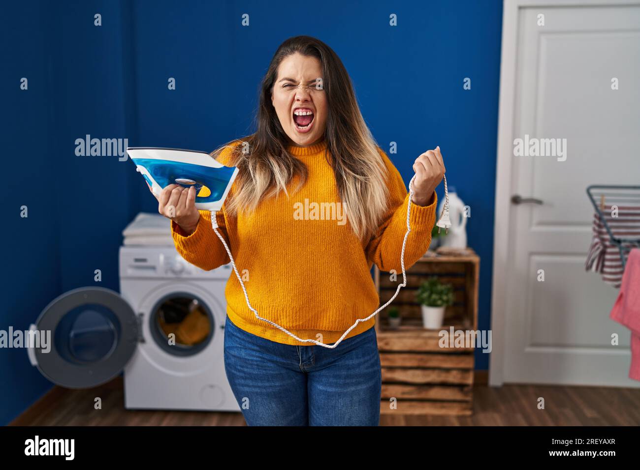 Young hispanic woman holding electric iron angry and mad screaming ...