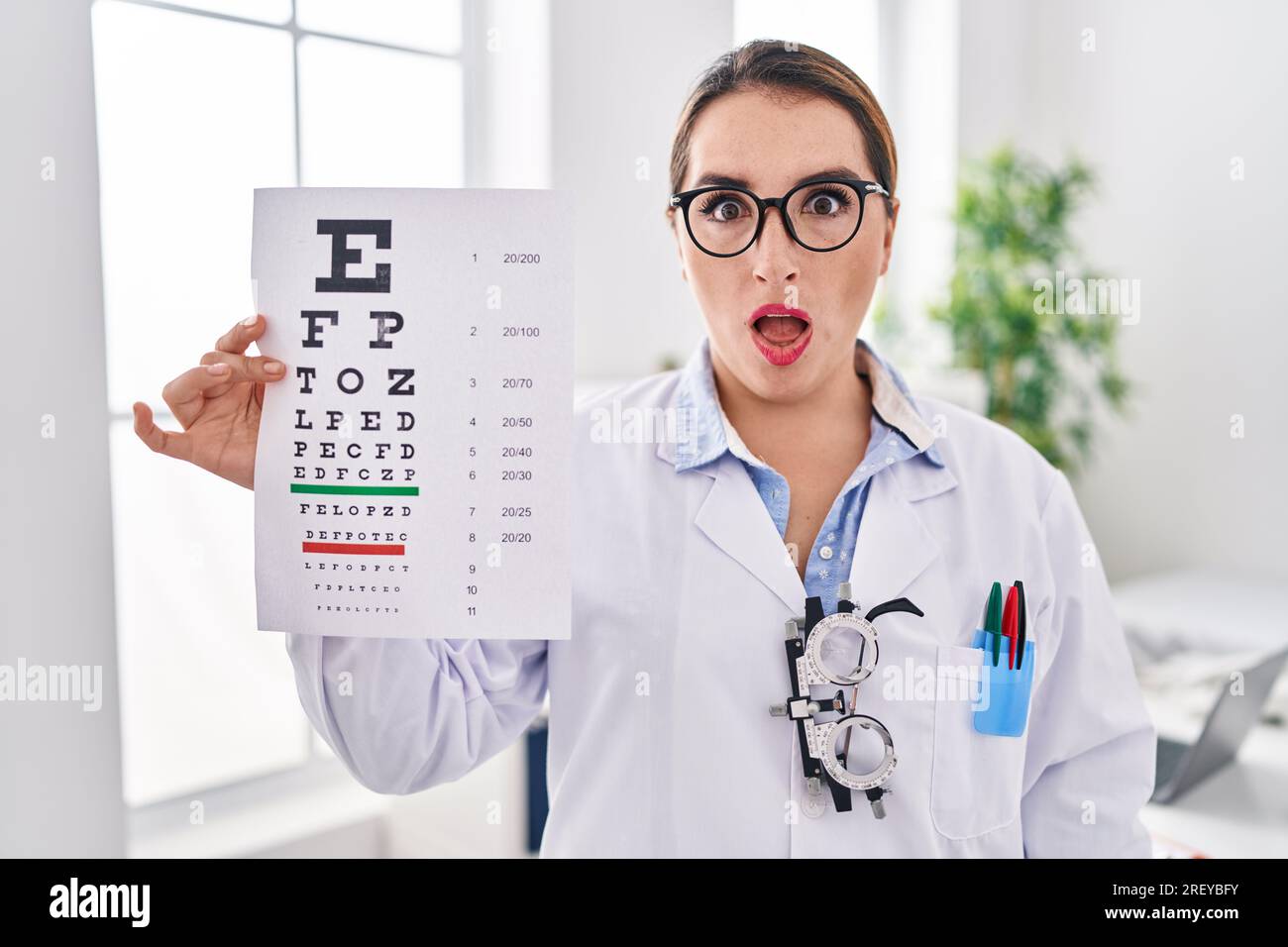 Young hispanic optician woman holding medical exam scared and amazed ...