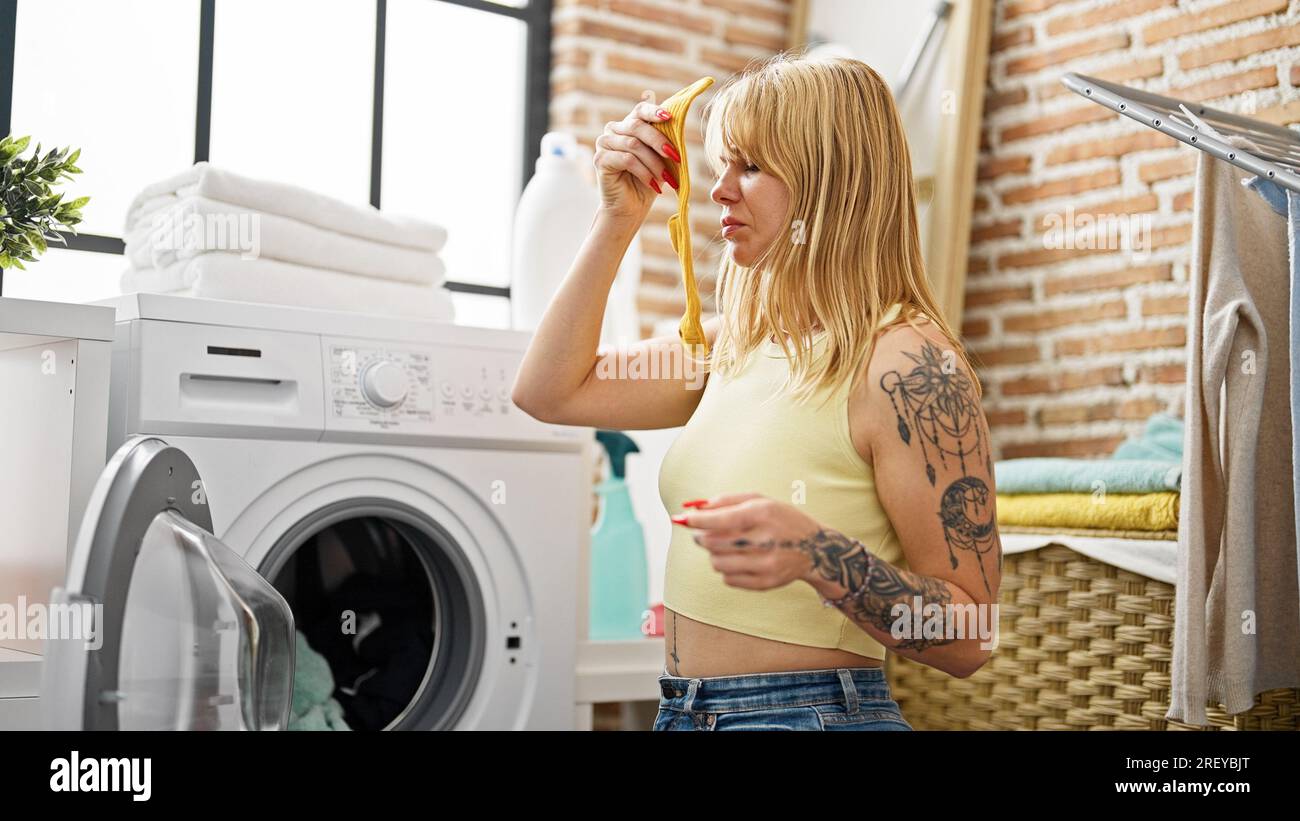 Young blonde woman washing clothes smelling dirty clothes at laundry ...