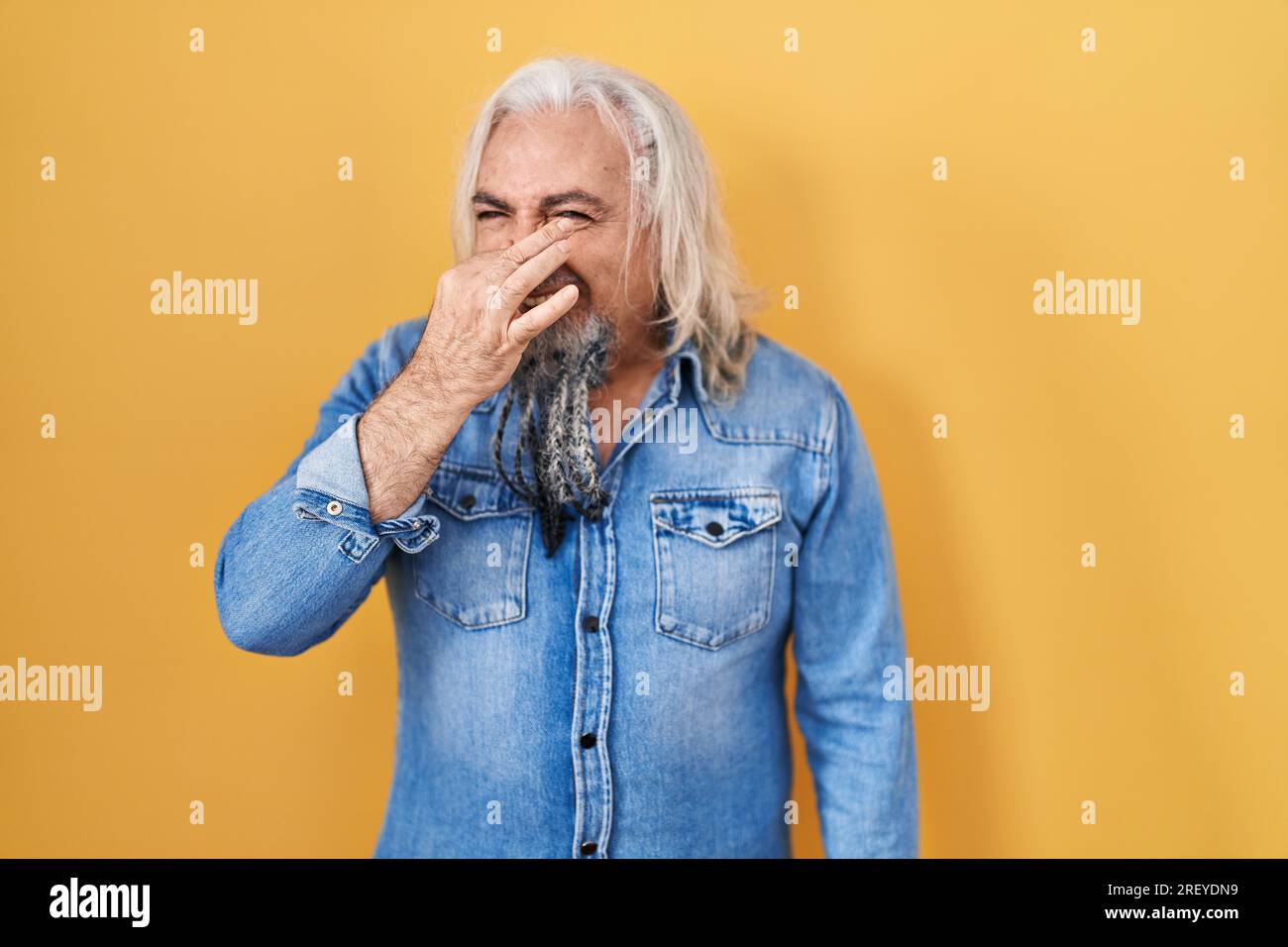 Middle age man with grey hair standing over yellow background smelling ...