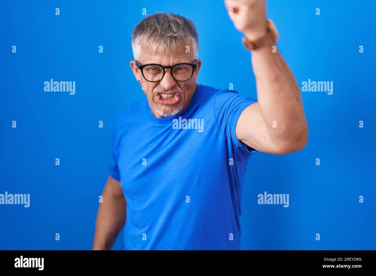 Hispanic man with grey hair standing over blue background angry and mad ...