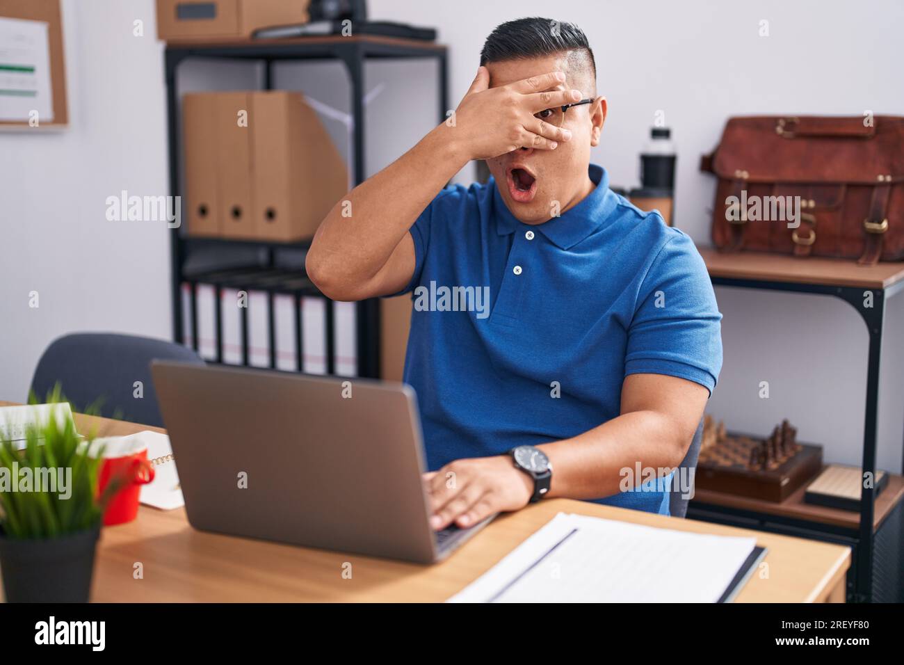 Young hispanic man working at the office with laptop peeking in shock ...