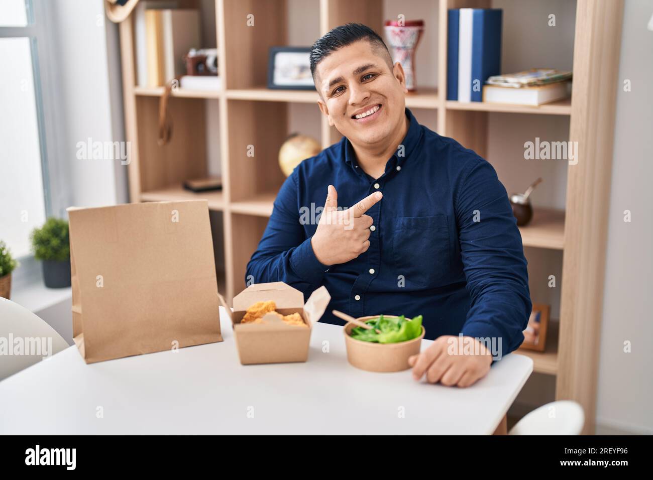 Hispanic young man eating take away food smiling happy pointing with ...