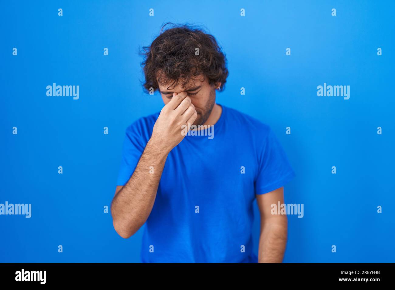 Hispanic young man standing over blue background tired rubbing nose and ...