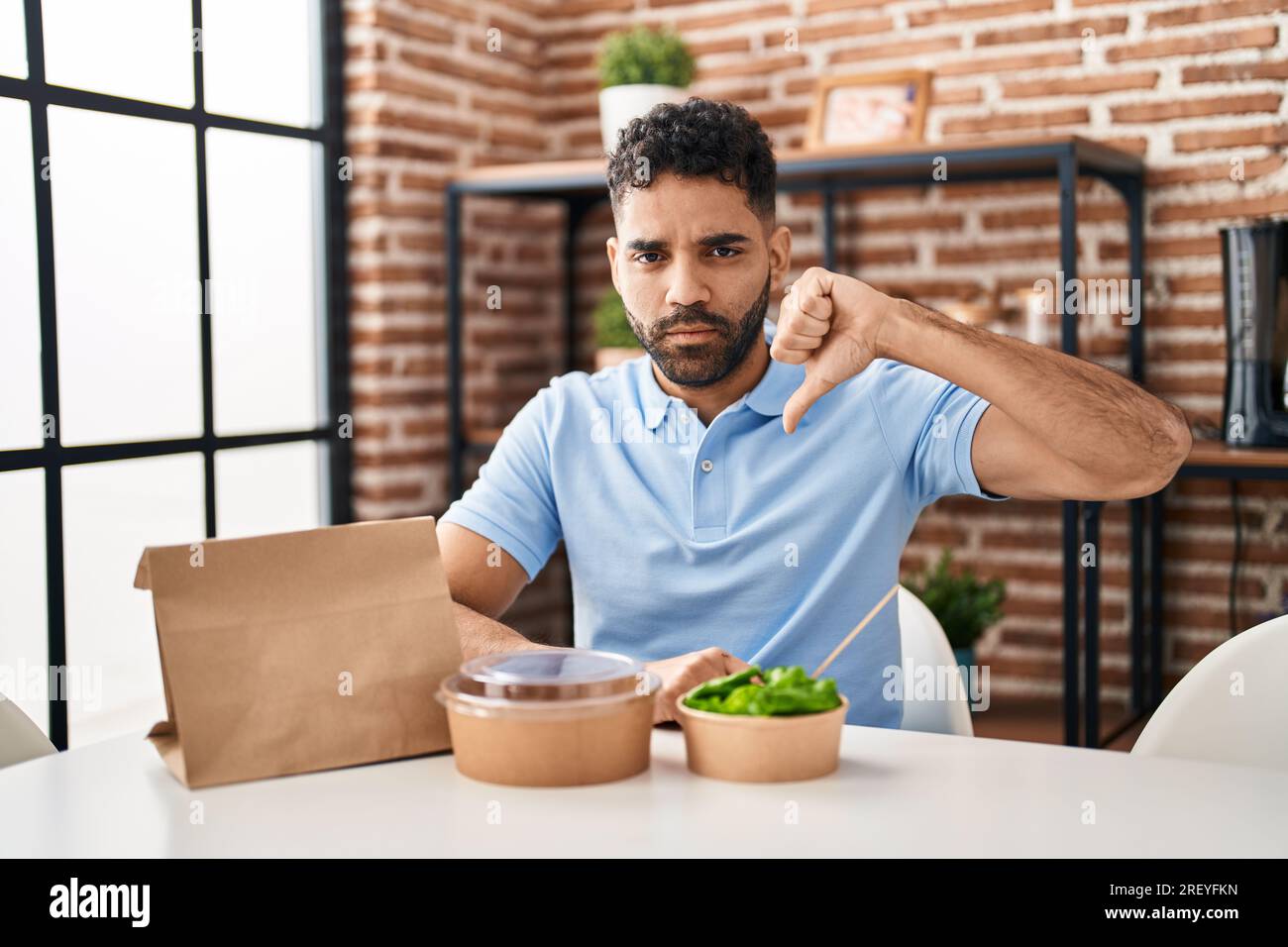 Hispanic man with beard eating delivery salad with angry face, negative ...
