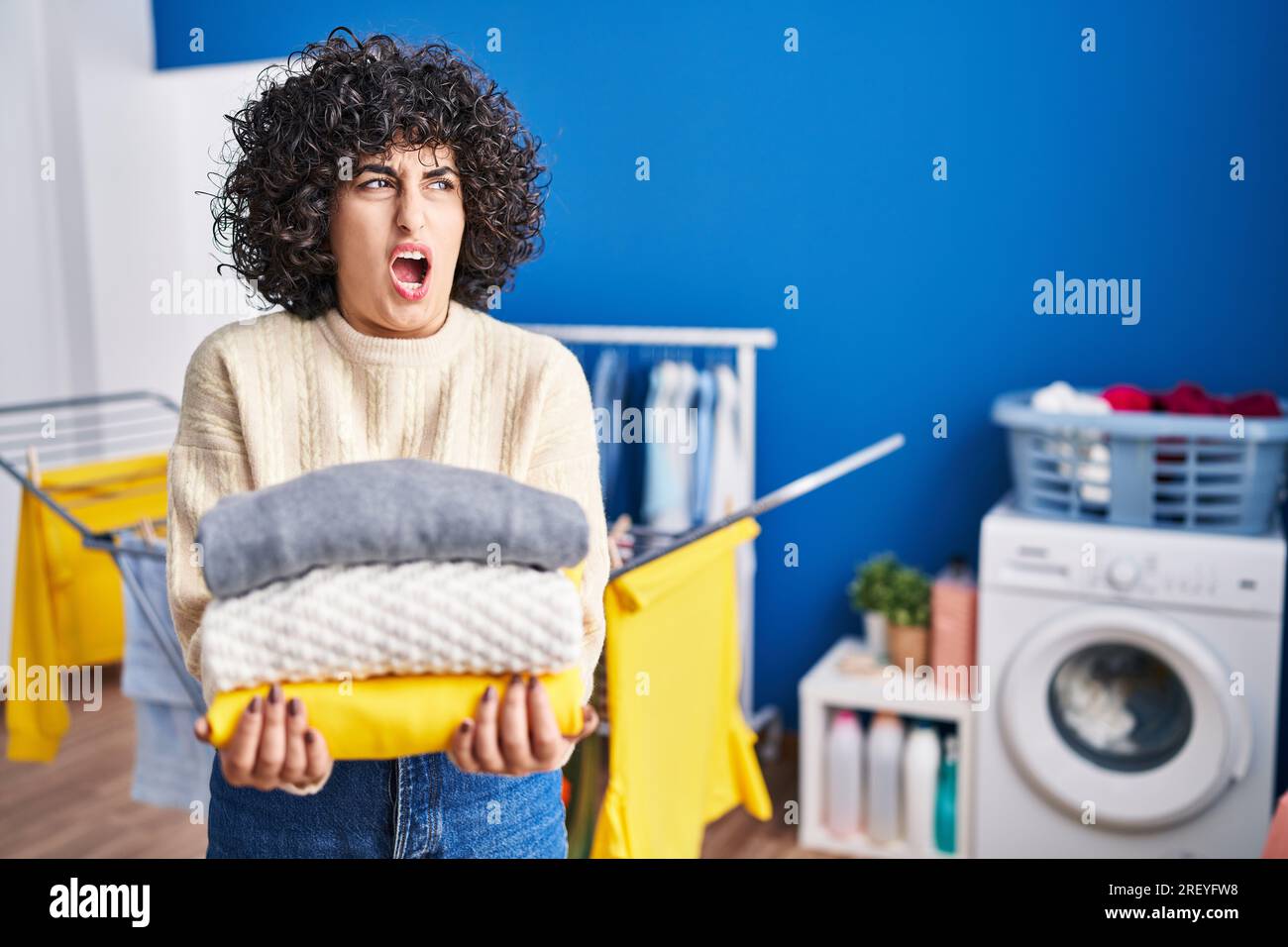 Young brunette woman with curly hair holding clean laundry angry and mad screaming frustrated ...