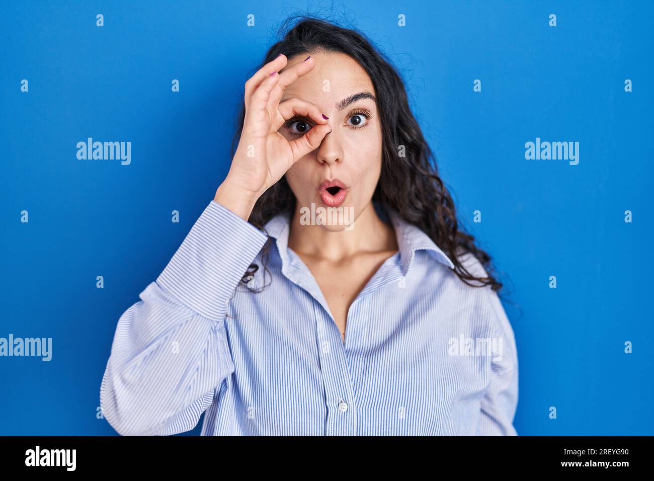 Young brunette woman standing over blue background doing ok gesture ...