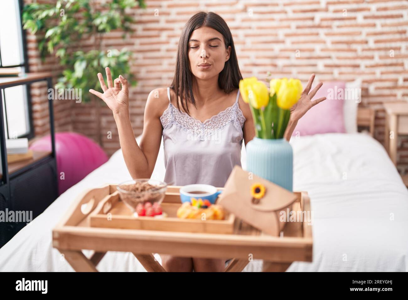 Brunette young woman eating breakfast sitting on the bed relax and smiling with eyes closed ...