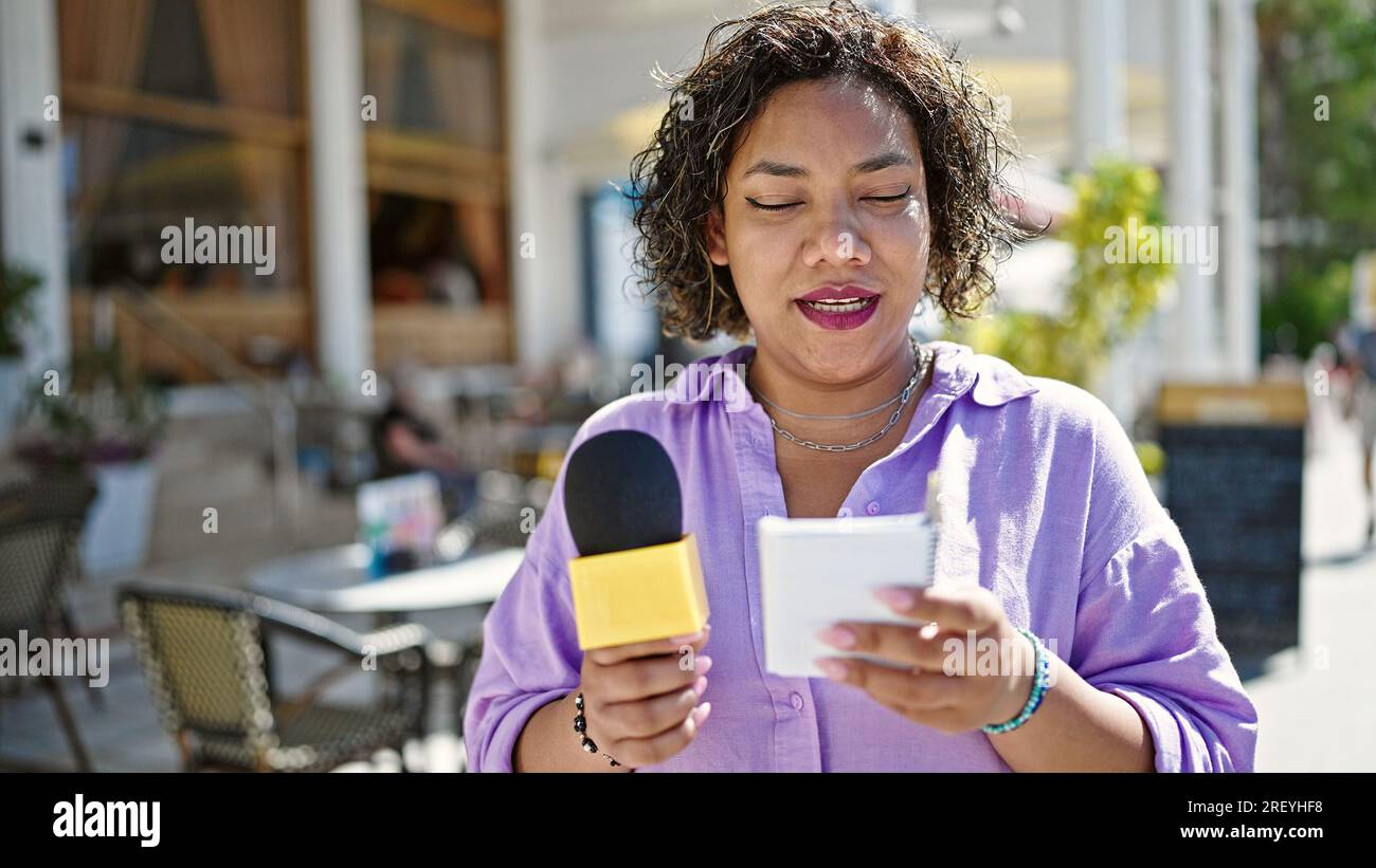 Young beautiful latin woman reporter working using microphone reading ...