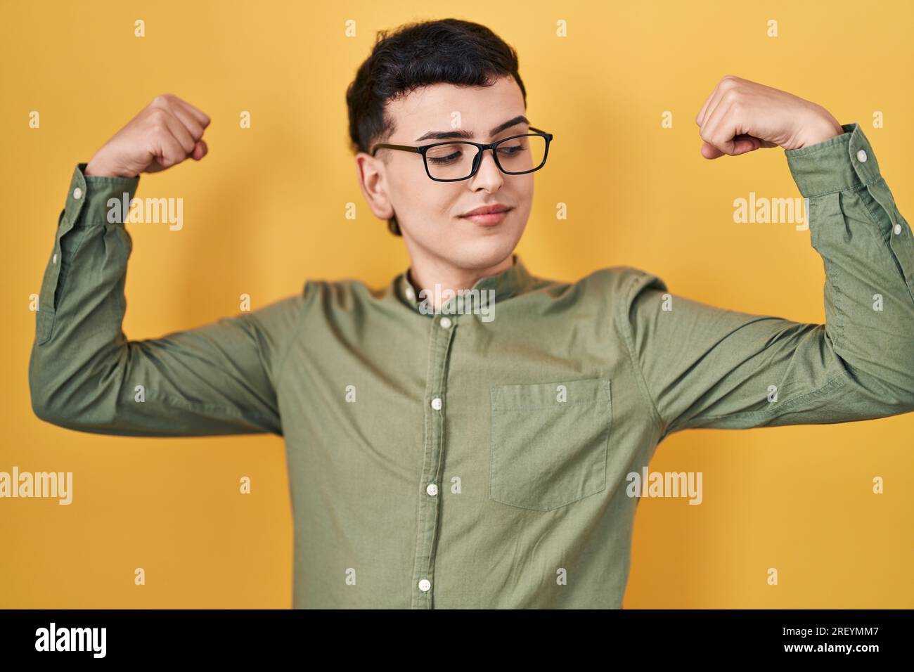 Non binary person standing over yellow background showing arms muscles ...