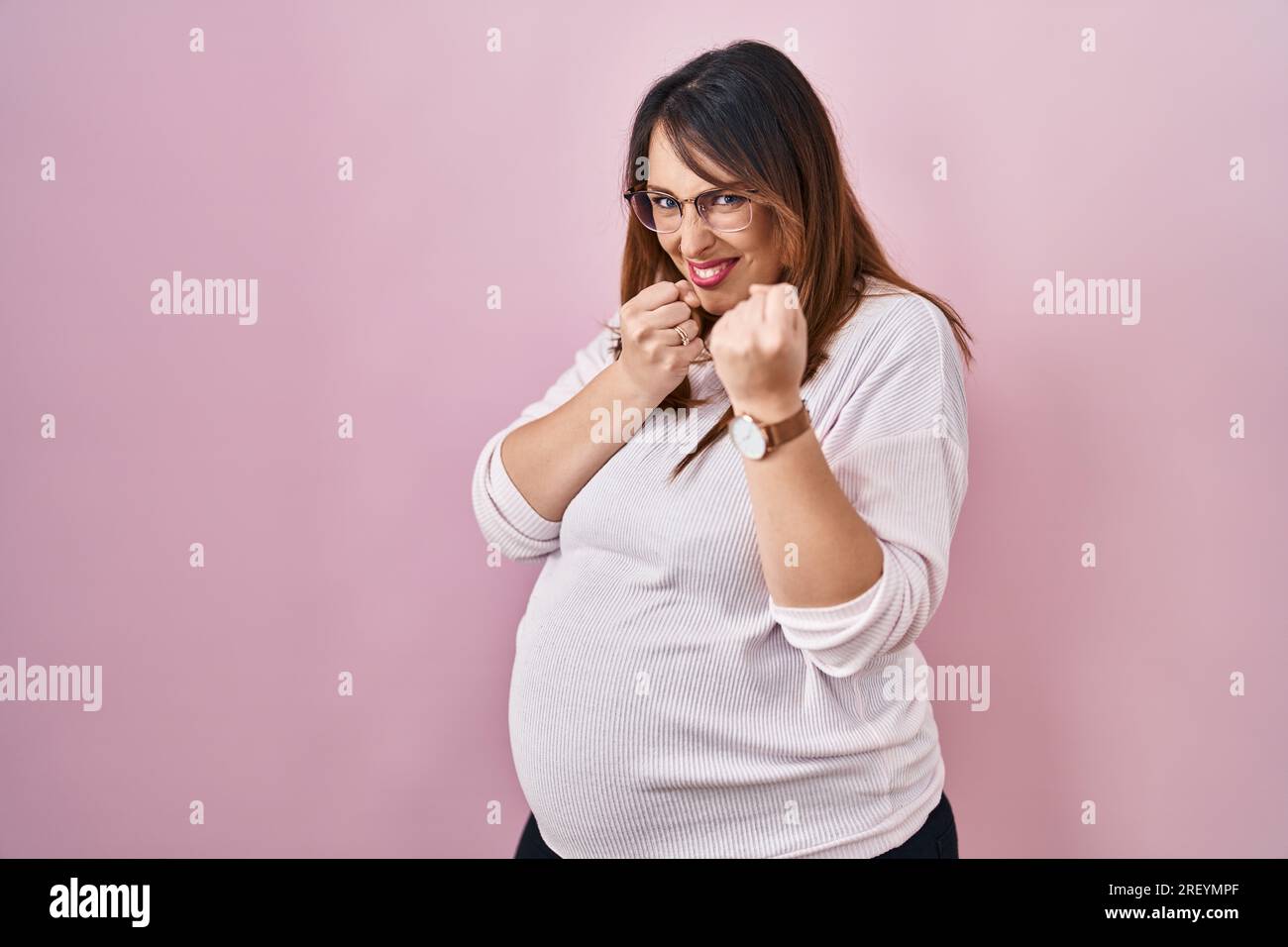 Pregnant woman standing over pink background ready to fight with fist ...