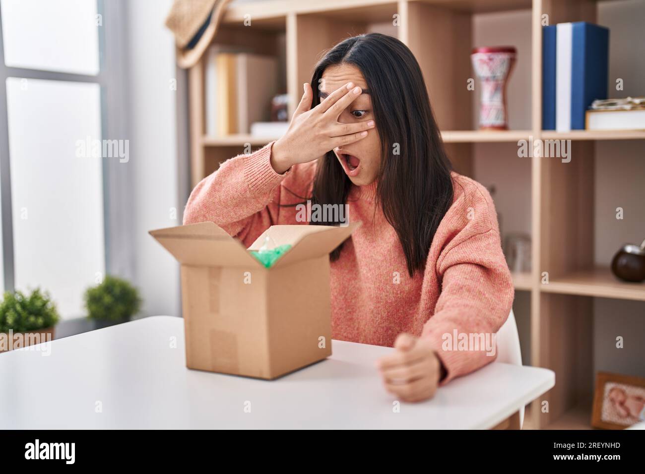 Young south asian woman opening cardboard box peeking in shock covering ...