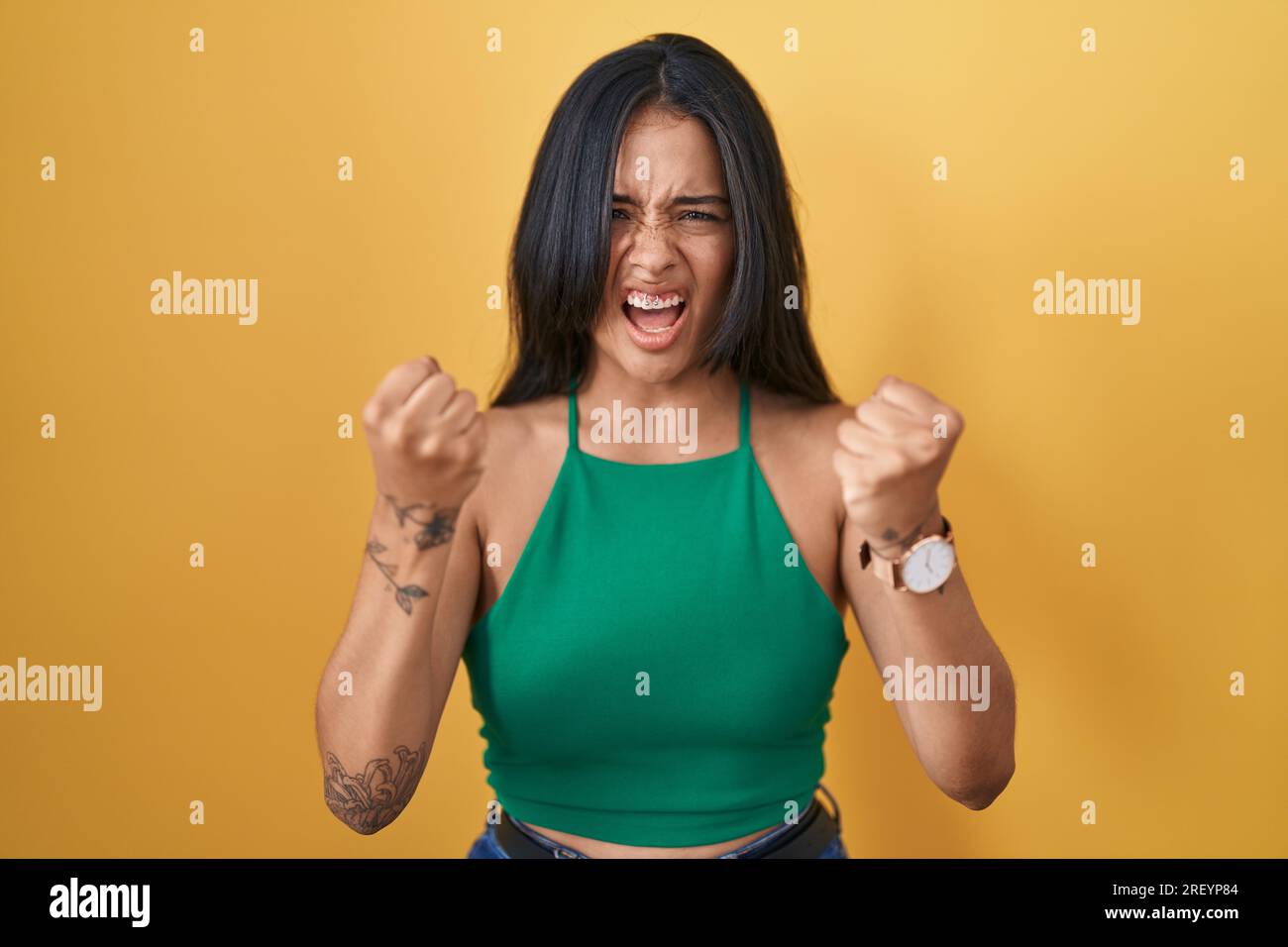 Brunette woman standing over yellow background angry and mad raising ...