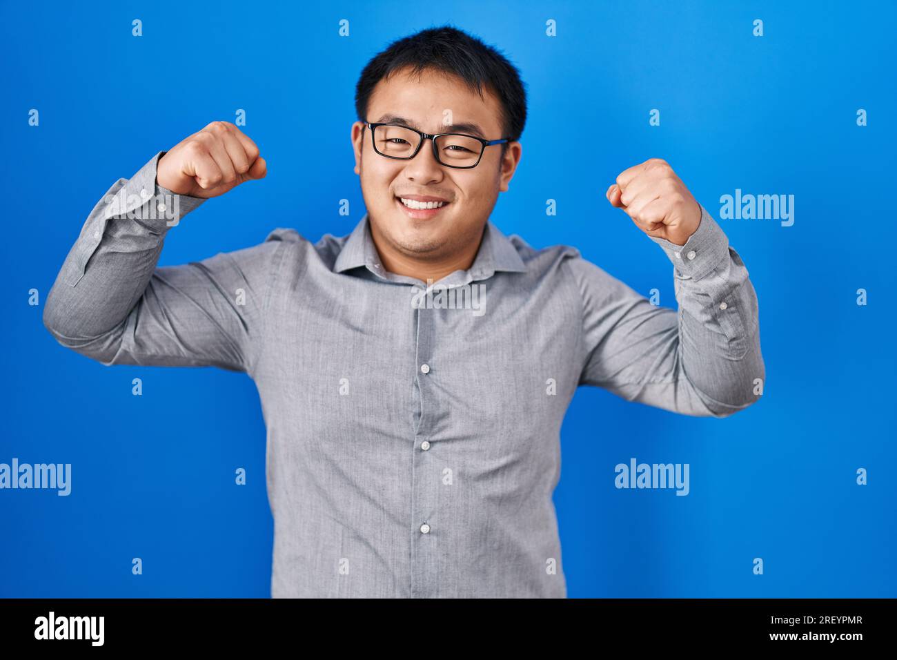 Young chinese man standing over blue background showing arms muscles ...