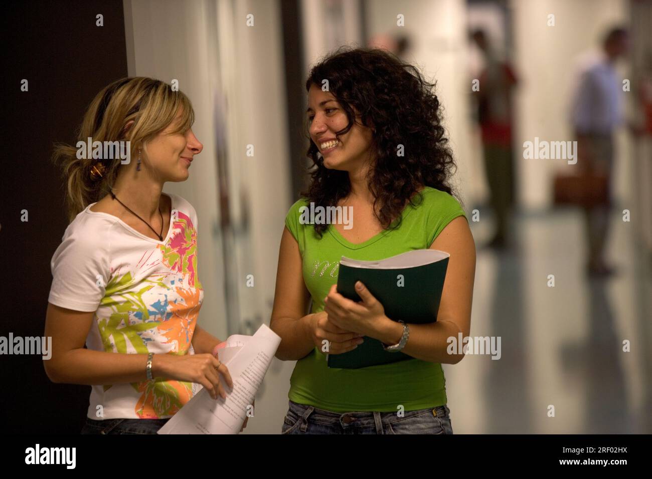 Two women conversing in an indoor hallway, holding papers and smiling ...