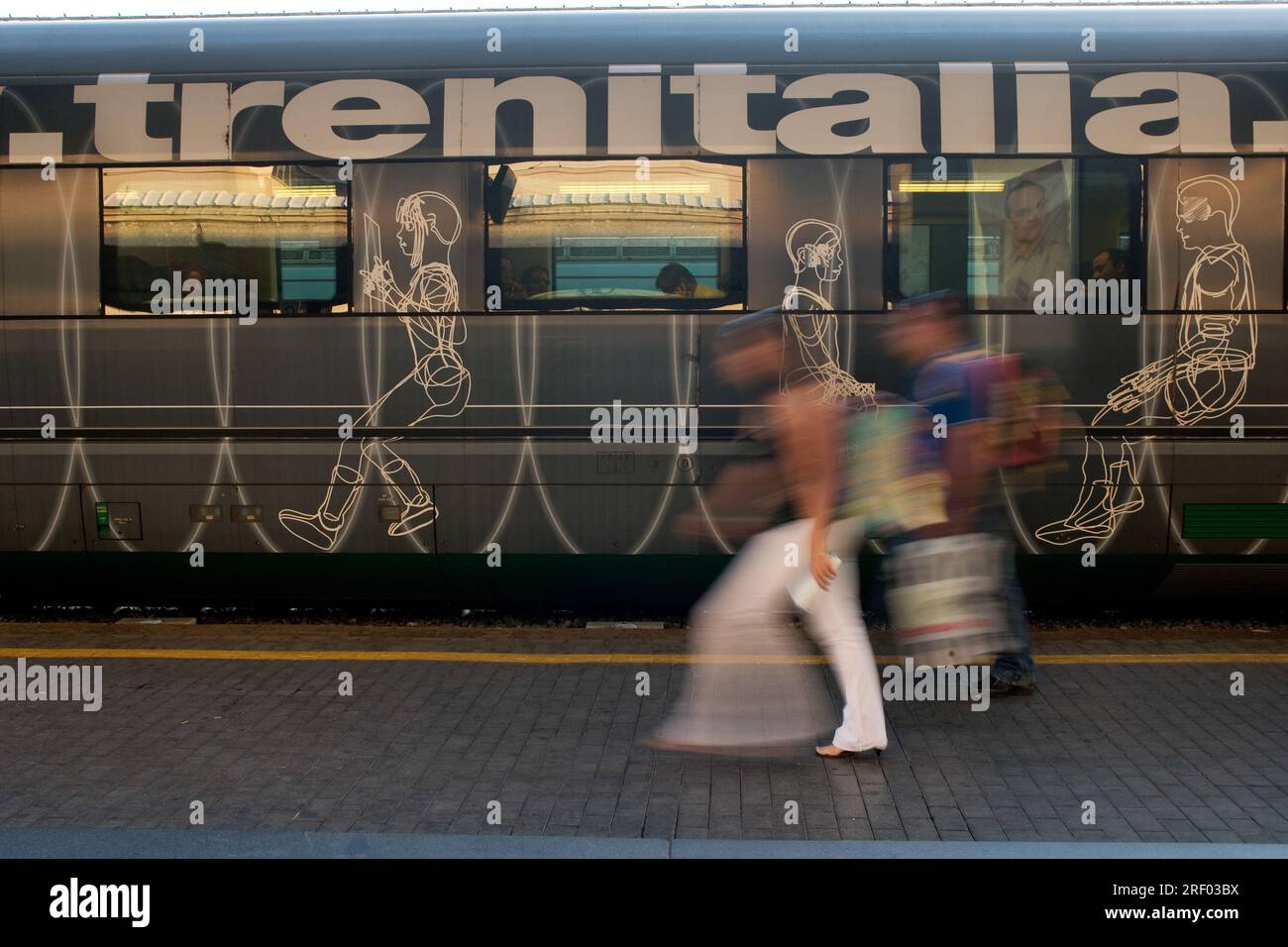 Passengers walk past a Trenitalia train at a platform, featuring ...