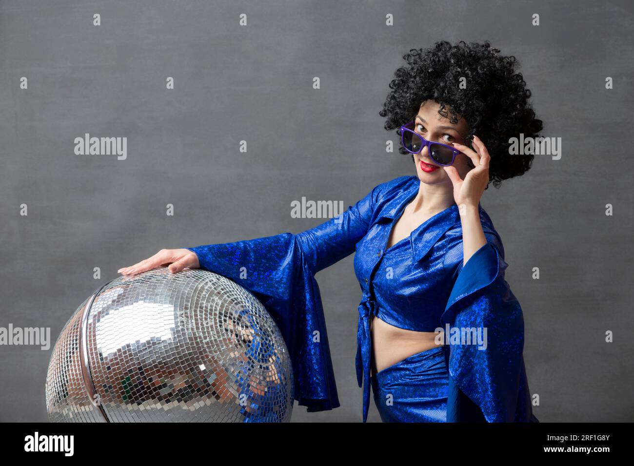 Girl with a disco ball in an afro wig on a gray background. Disco style ...