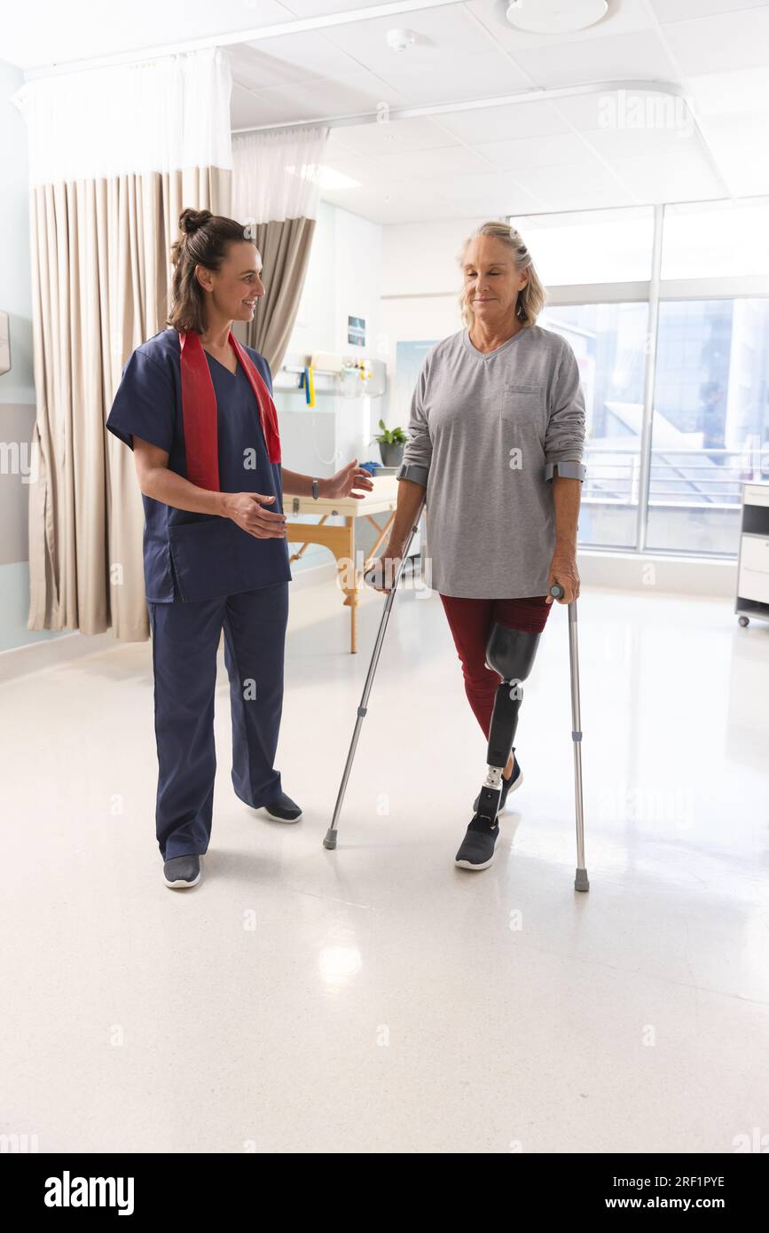 Caucasian female physiotherapist and senior woman with artificial leg walking with crutches ...