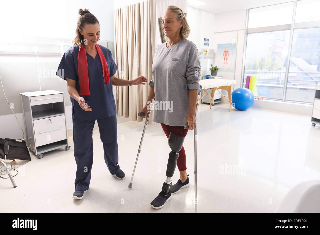 Caucasian female physiotherapist and senior woman with artificial leg walking with crutches ...