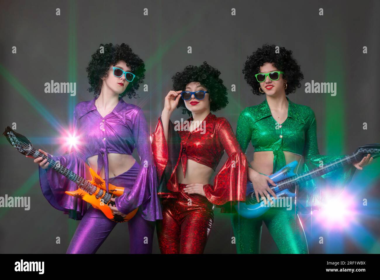 Beautiful girls dressed in disco style in afro wigs pose with guitars in the rays of stage light ...