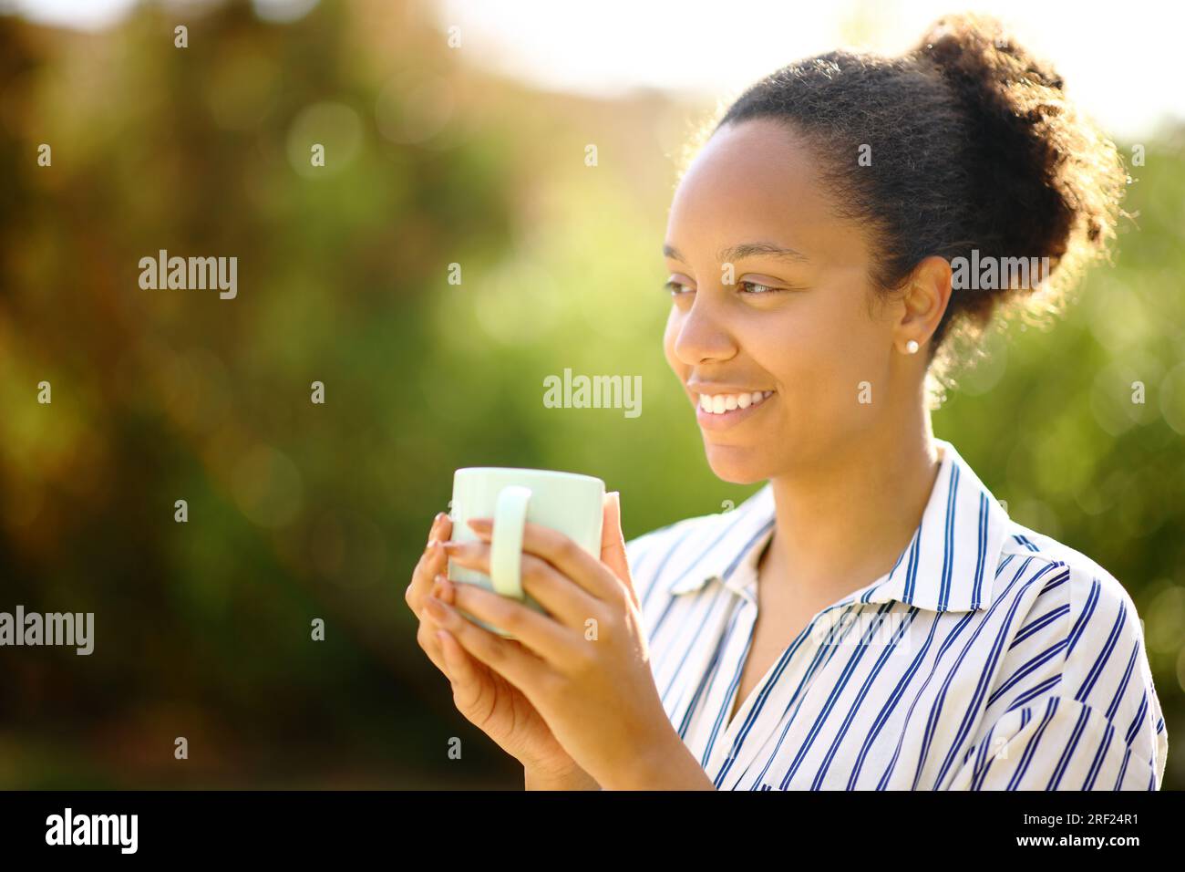 Happy black woman holding coffee mug contemplating views in a park ...