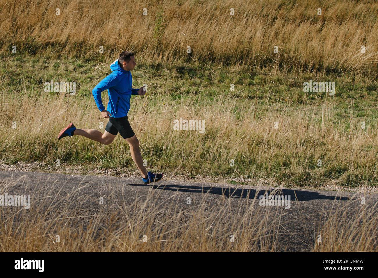 middle-aged male runner in blue jacket running road in field of dry ...