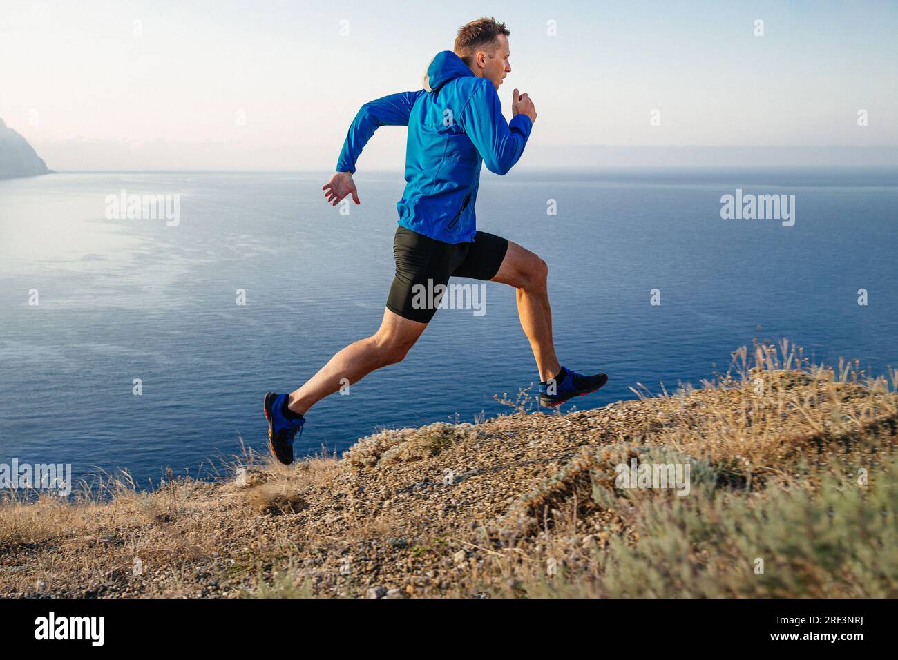 male runner running uphill trail on edge in blue jacket and black ...