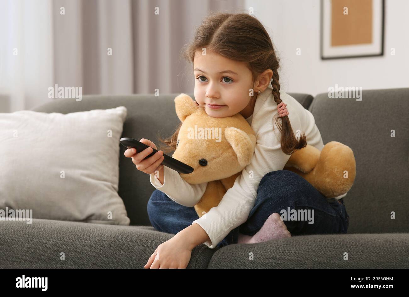 Little girl changing TV channels with remote control on sofa at home ...