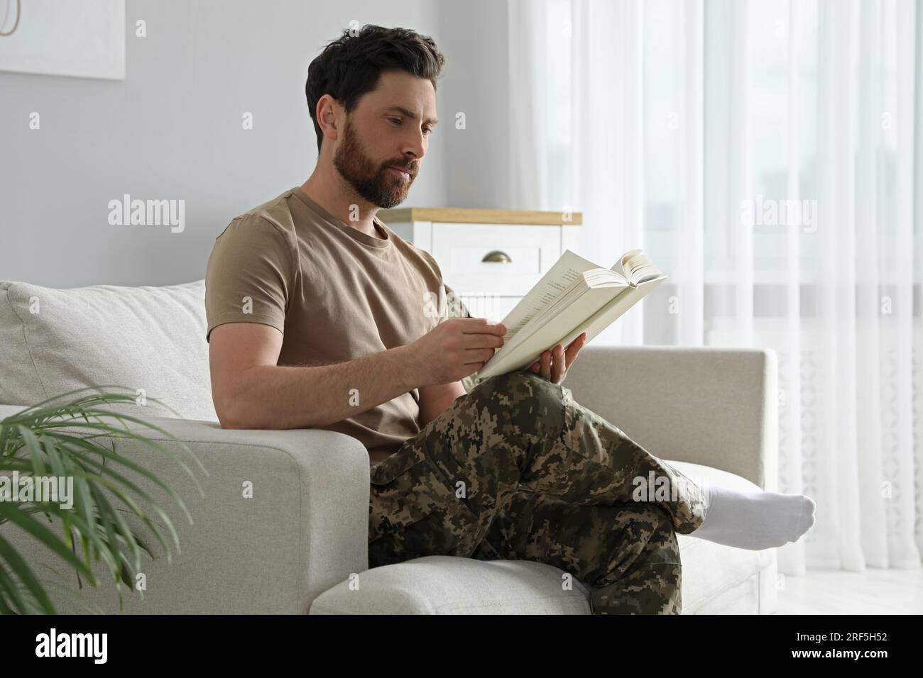 Soldier reading book on soft sofa in living room. Military service ...