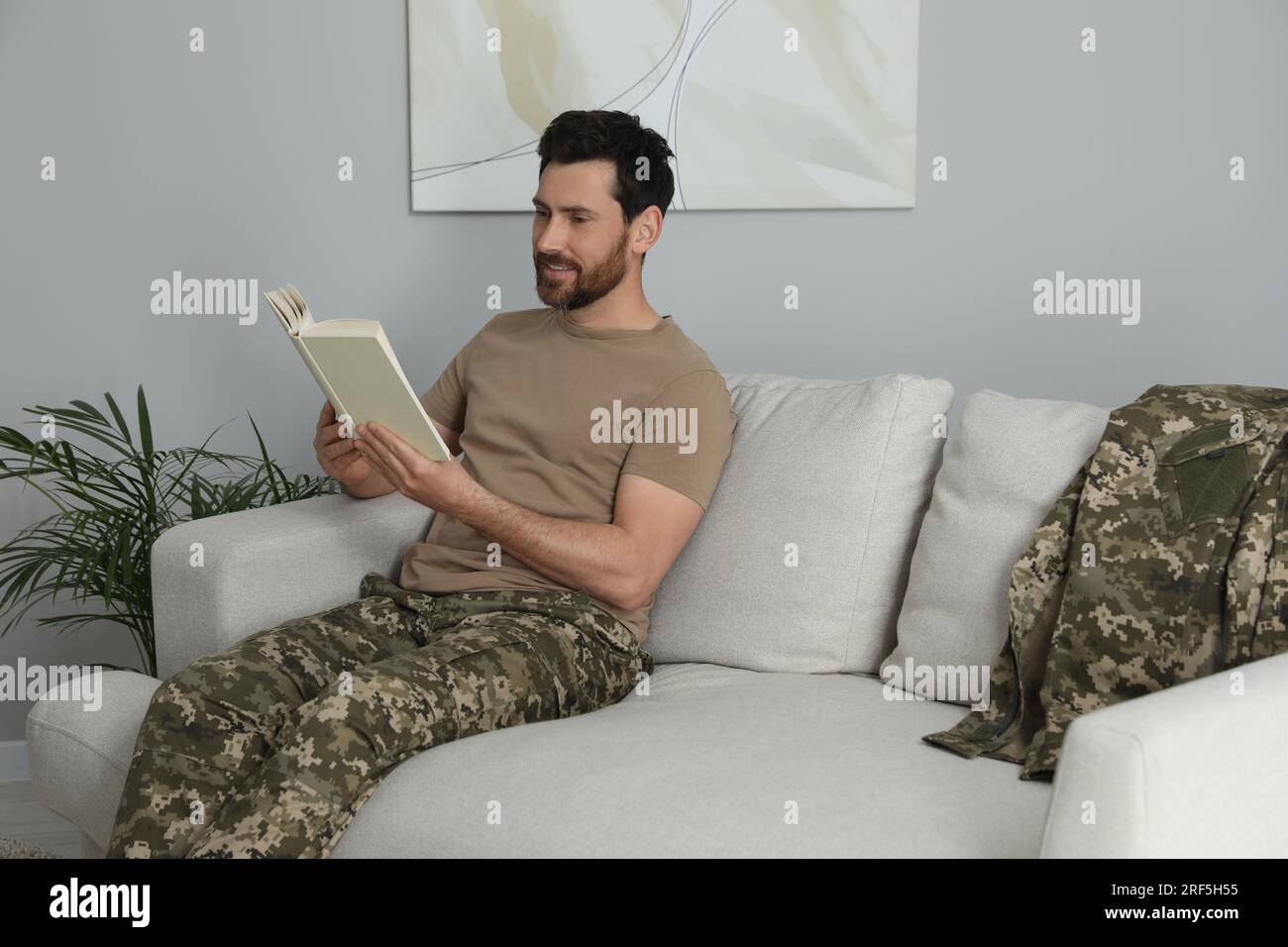 Soldier reading book on soft sofa in living room. Military service ...