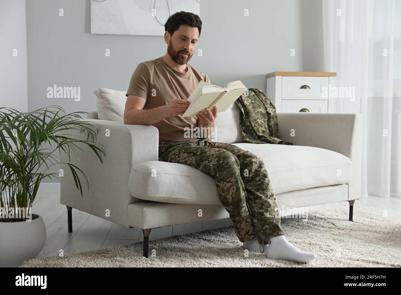 Soldier reading book on soft sofa in living room. Military service ...