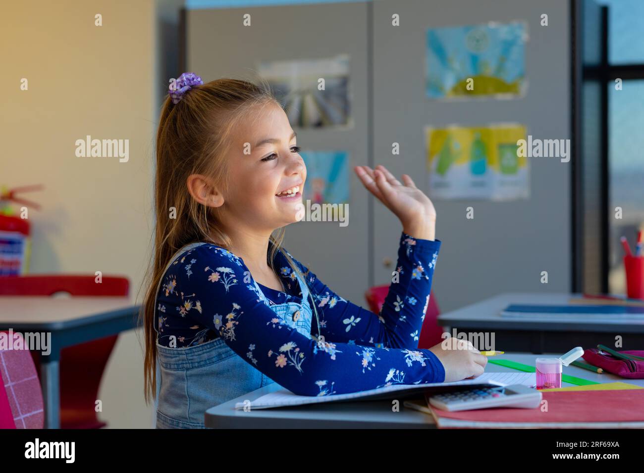 Happy caucasian schoolgirl sitting at desk and waving hand in classroom ...