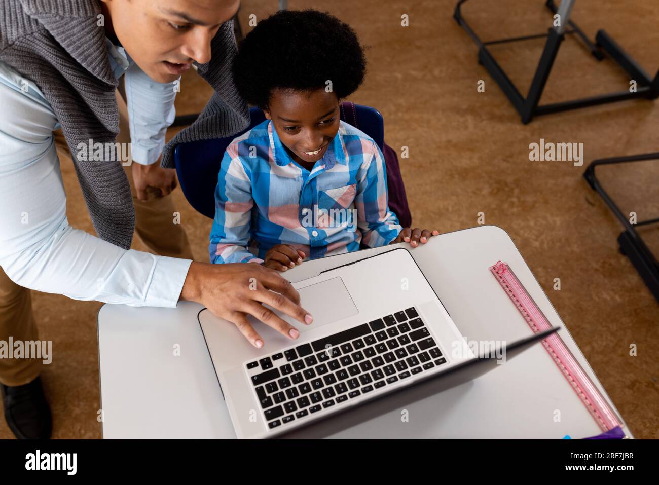 Diverse male teacher using laptop teaching boy at desk in elementary ...