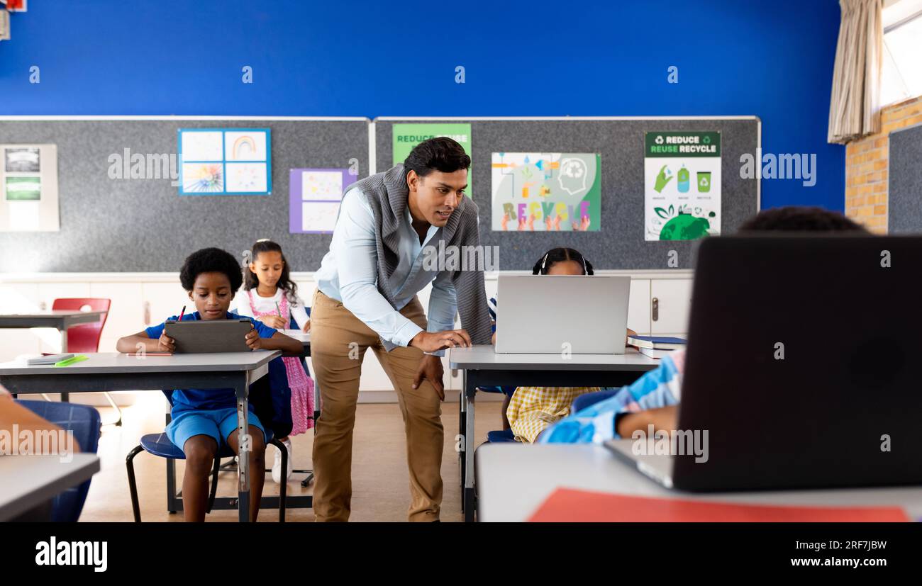 Diverse male teacher using laptop teaching girl in class at elementary ...