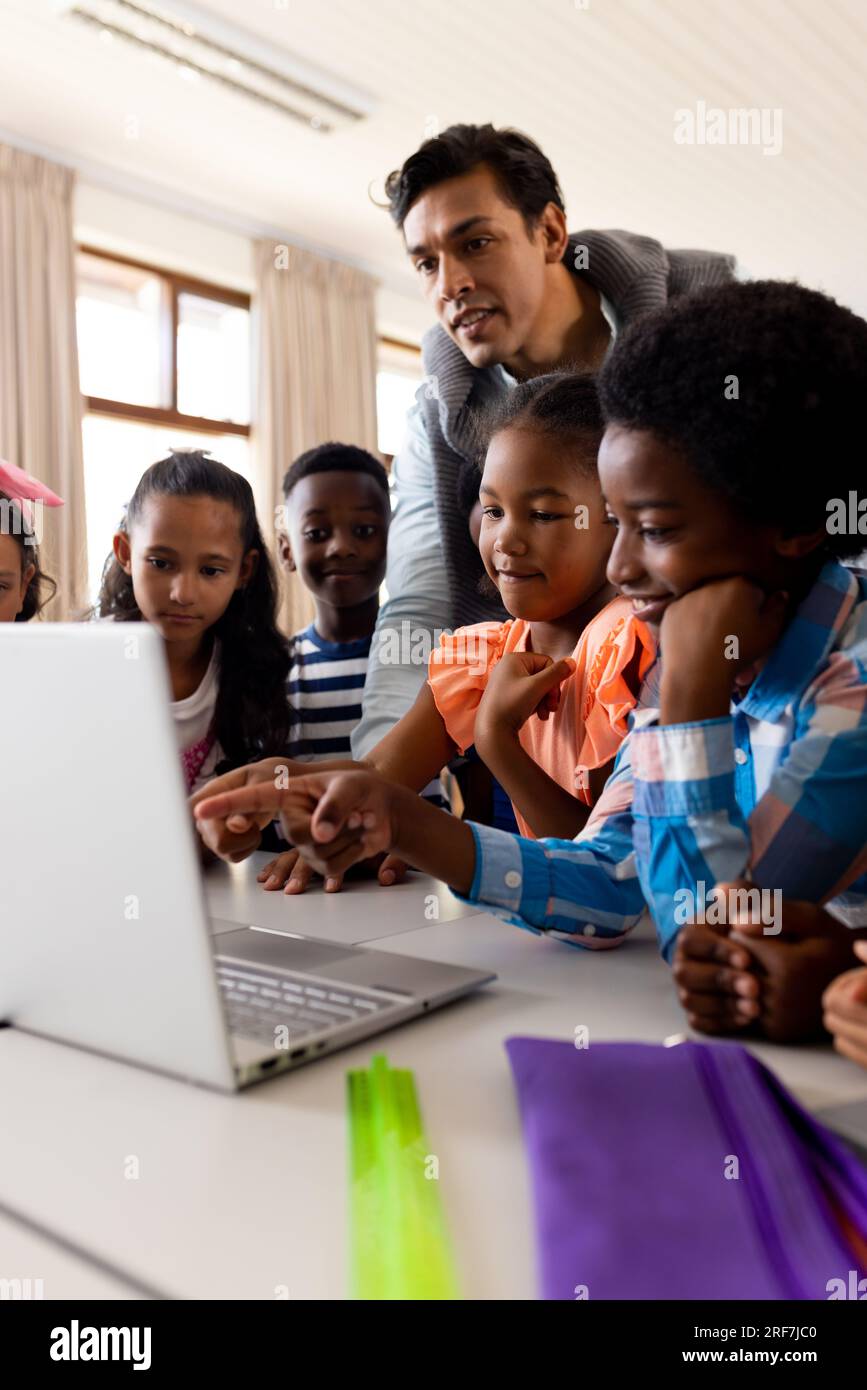 Diverse male teacher using laptop teaching children in class at ...