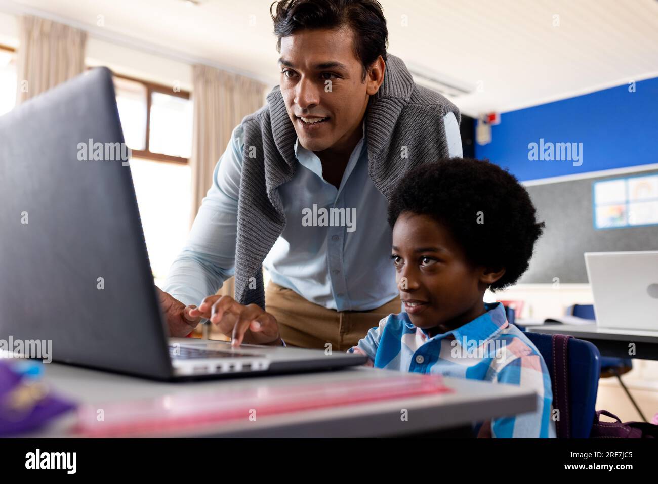 Diverse male teacher using laptop teaching boy in class at elementary ...