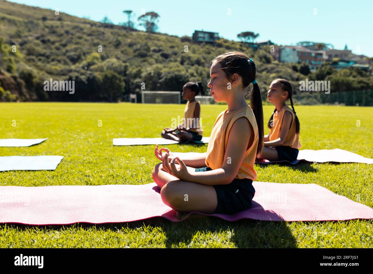 Diverse schoolgirls practicing yoga and meditating in sports field at ...