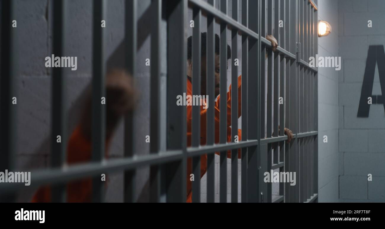 African American prisoner in orange uniform leans on jail cell bars ...