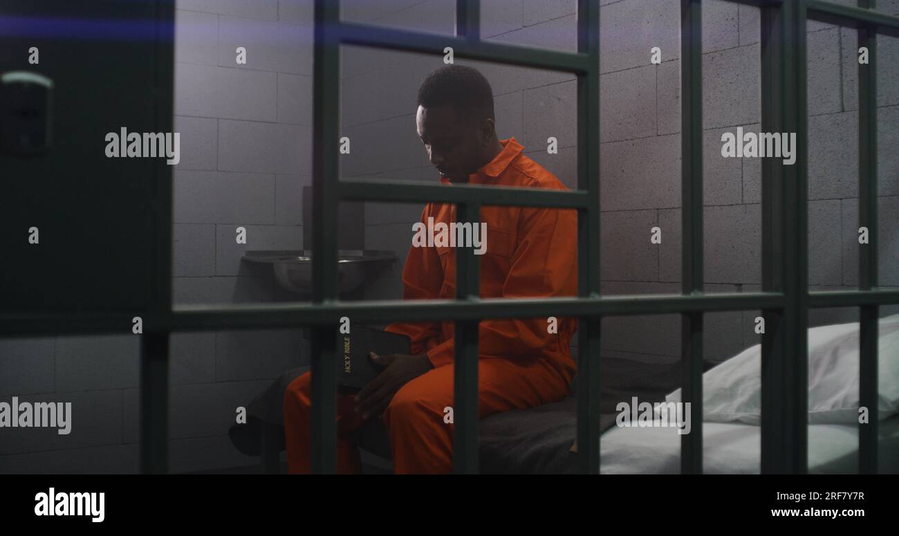 African American prisoner in orange uniform sits on the bed behind bars ...